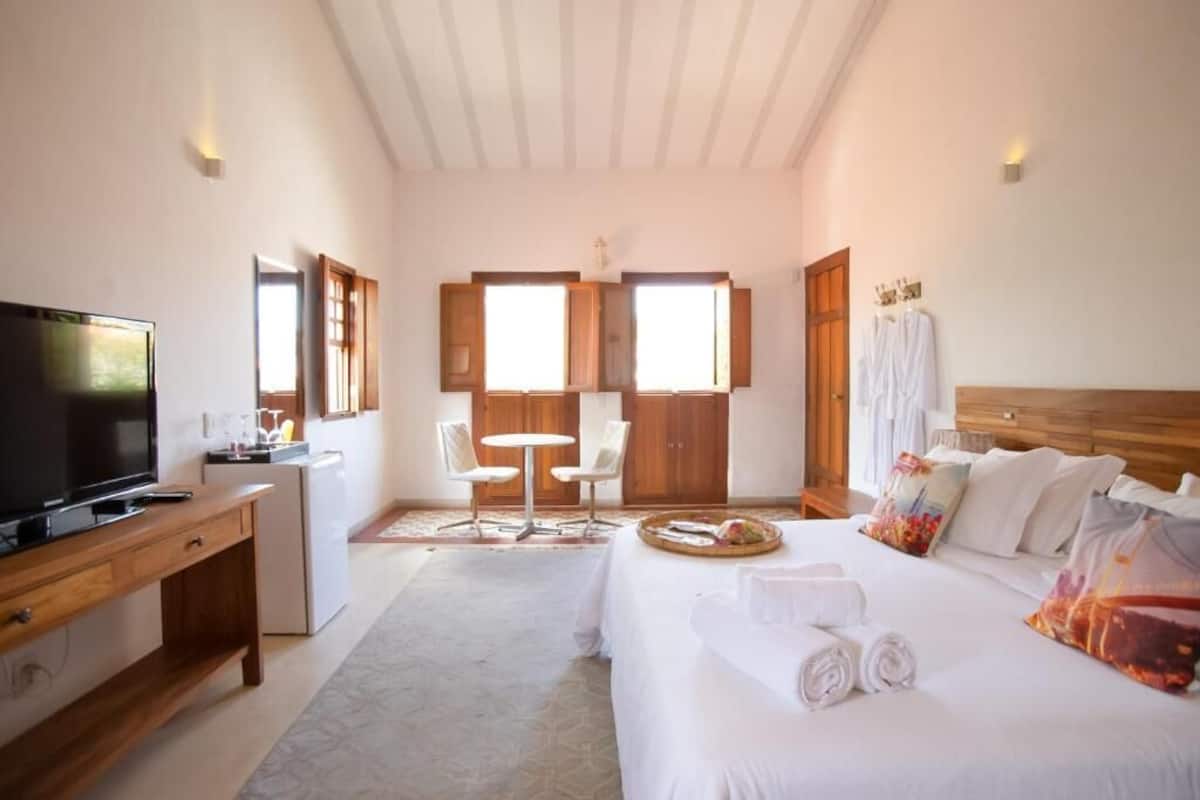 Quarto de hotel com cama, televisão, escrivaninha, cadeira, mesinha de apoio e guarda-roupa.