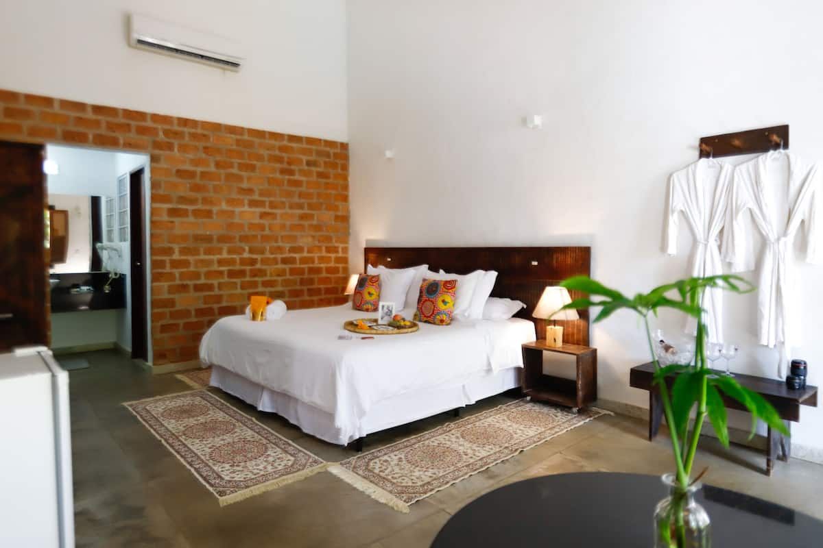 Quarto de hotel com parede de tijolos, cama com roupa de cama branca, mesa de cabeceira com abajur, pequena mesa com um vaso e um espelho.