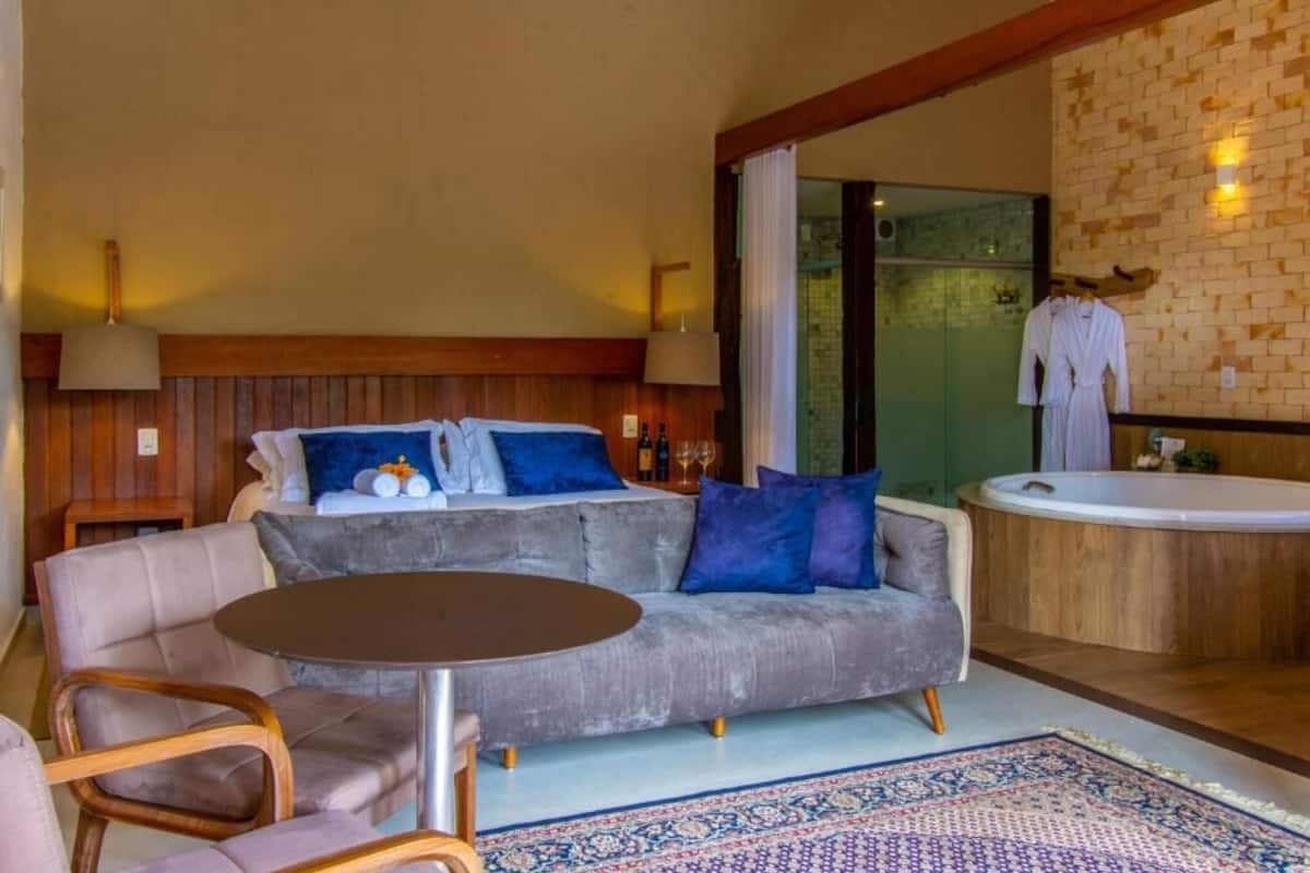 Quarto de hotel moderno com sofá cinza, mesa redonda, banheira grande e parede de madeira.