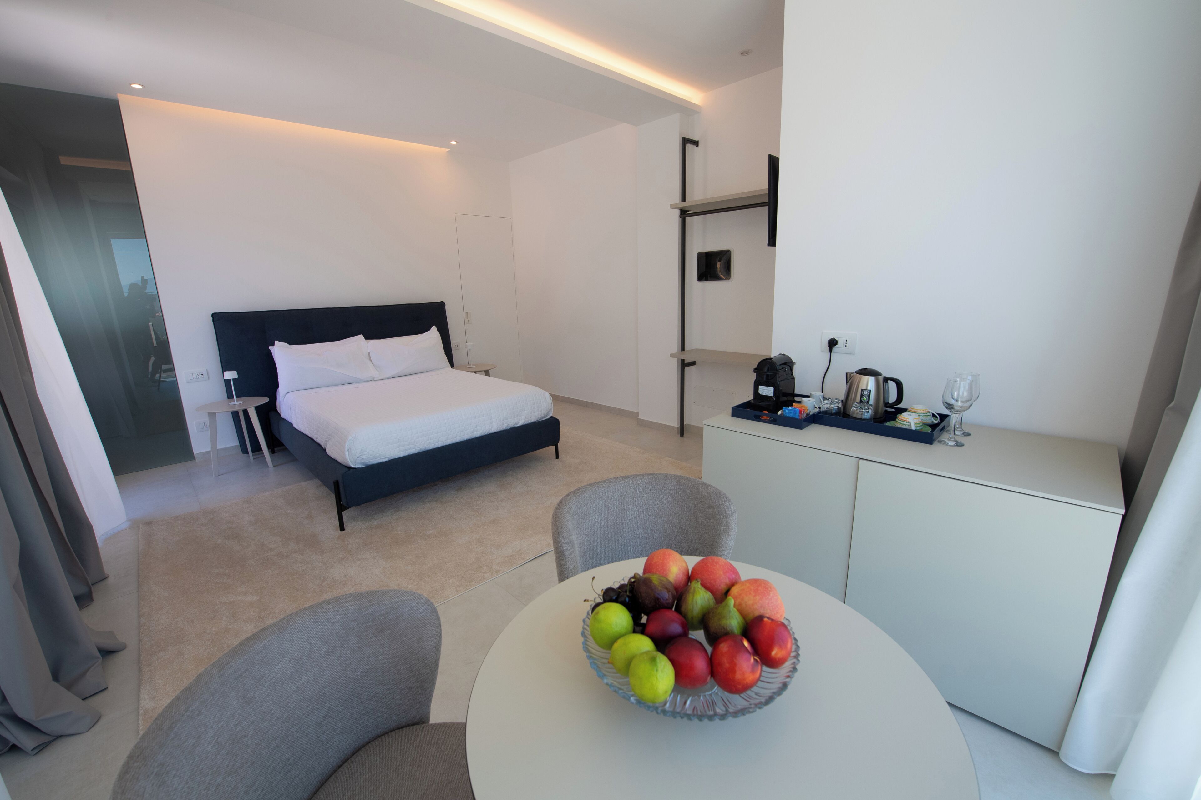 Quarto de hotel moderno com cama, mesa de jantar com frutas, cozinha compacta e área de estar.