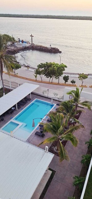 Piscina com espreguiçadeiras e palmeiras, área de praia com uma cruz, e vista para o mar.