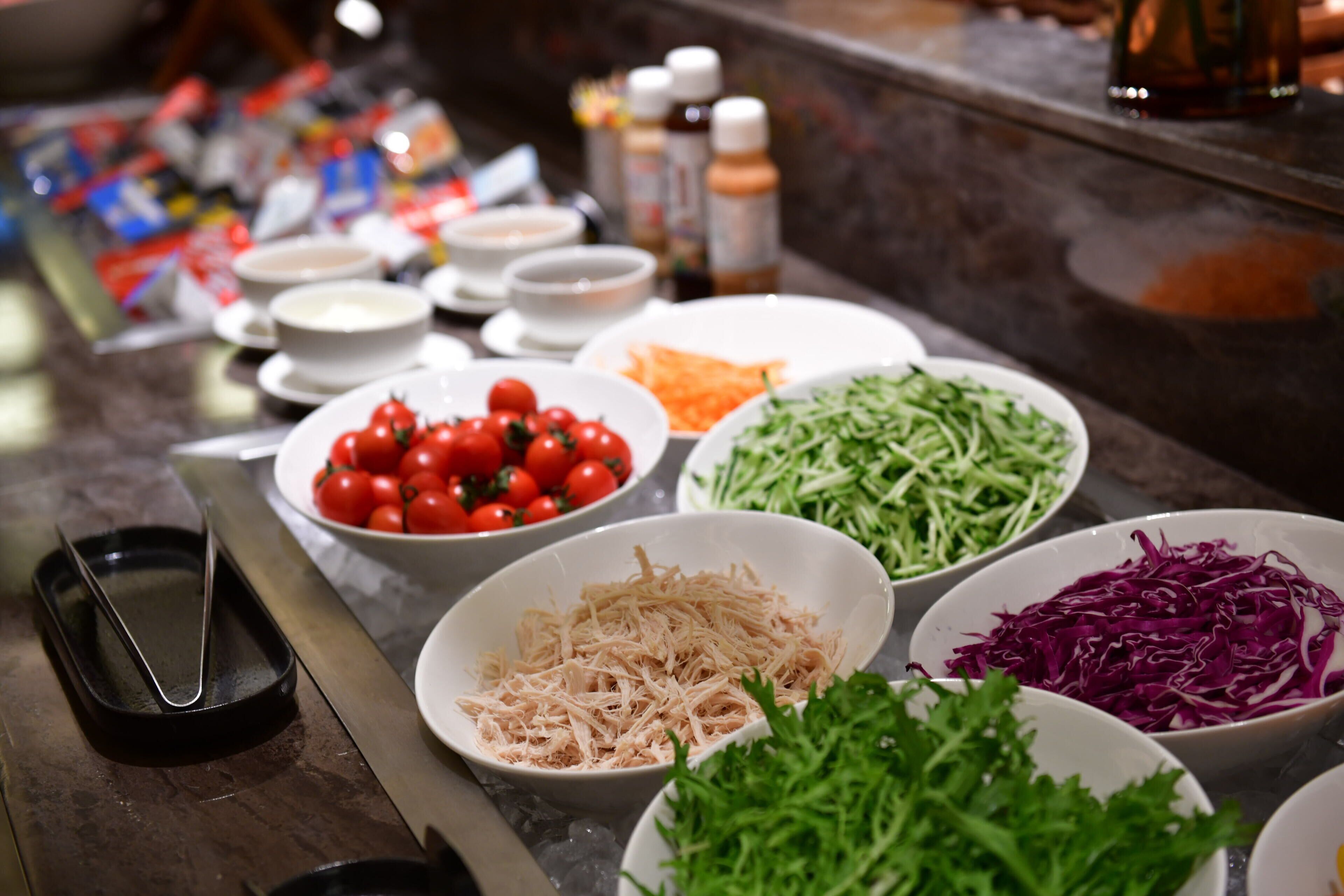 Mesa de buffet com diversos alimentos, incluindo tomates, frango desfiado e folhas verdes.