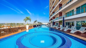 Piscina com espreguiçadeiras e palmeiras, cercada por um edifício com varandas.