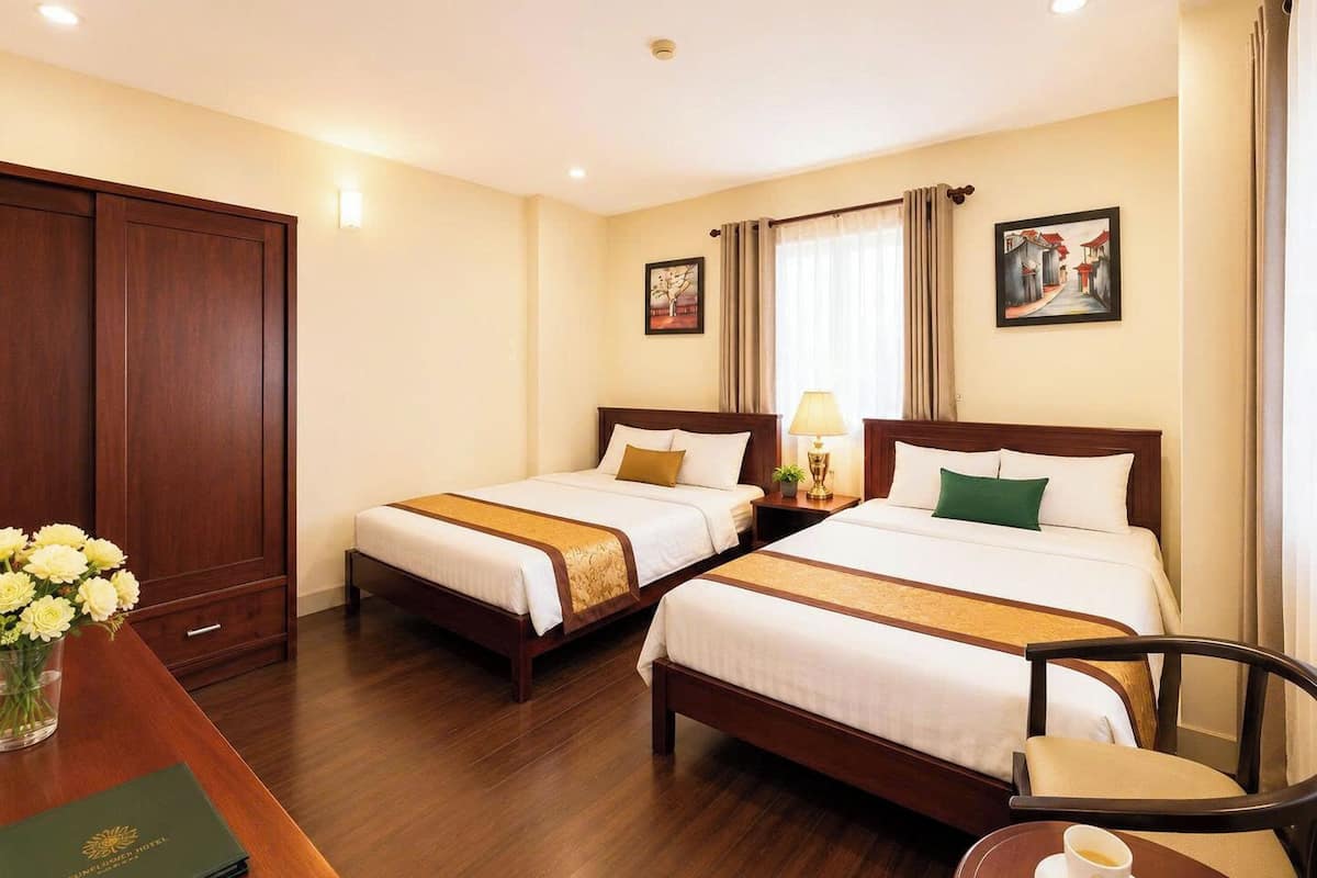 Quarto de hotel com duas camas, uma escrivaninha com cadeira, mesa de centro, um vaso de flores e um quadro na parede.