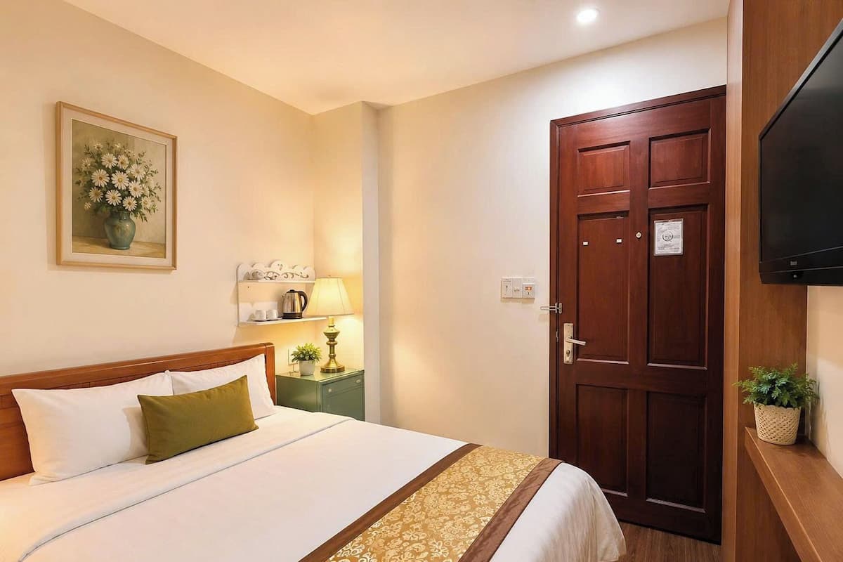 Quarto de hotel com cama, porta de madeira, televisão, pintura e mesa de cabeceira com abajur e planta.