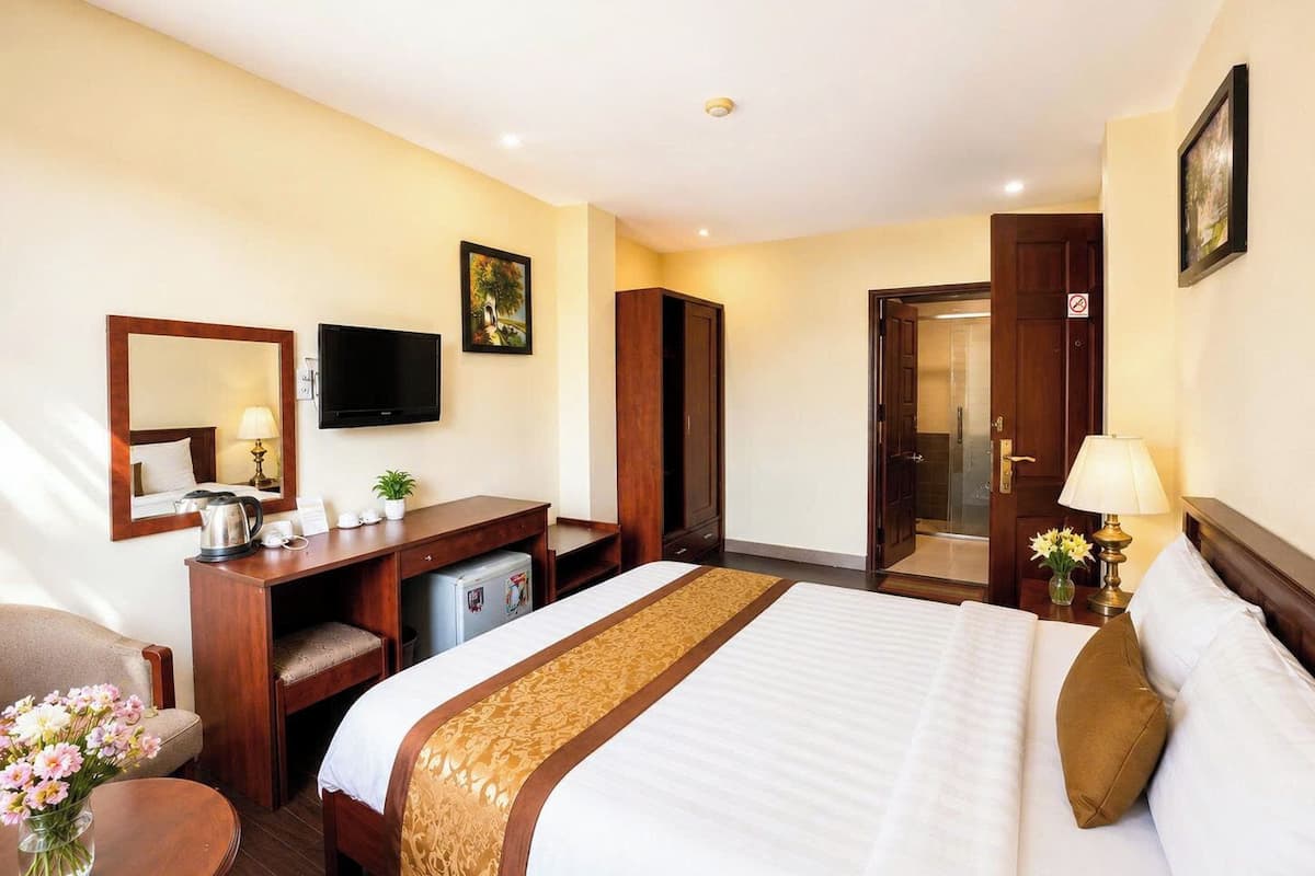 Quarto de hotel com cama, secretária com computador, televisão, espelho, porta e uma pequena mesa com flores.