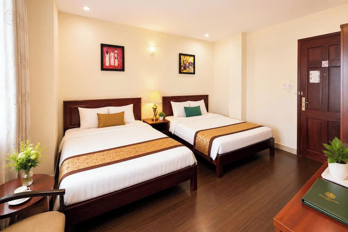 Quarto de hotel com duas camas, piso de madeira, escrivaninha com cadeira, mesinha com vaso e porta com placa.