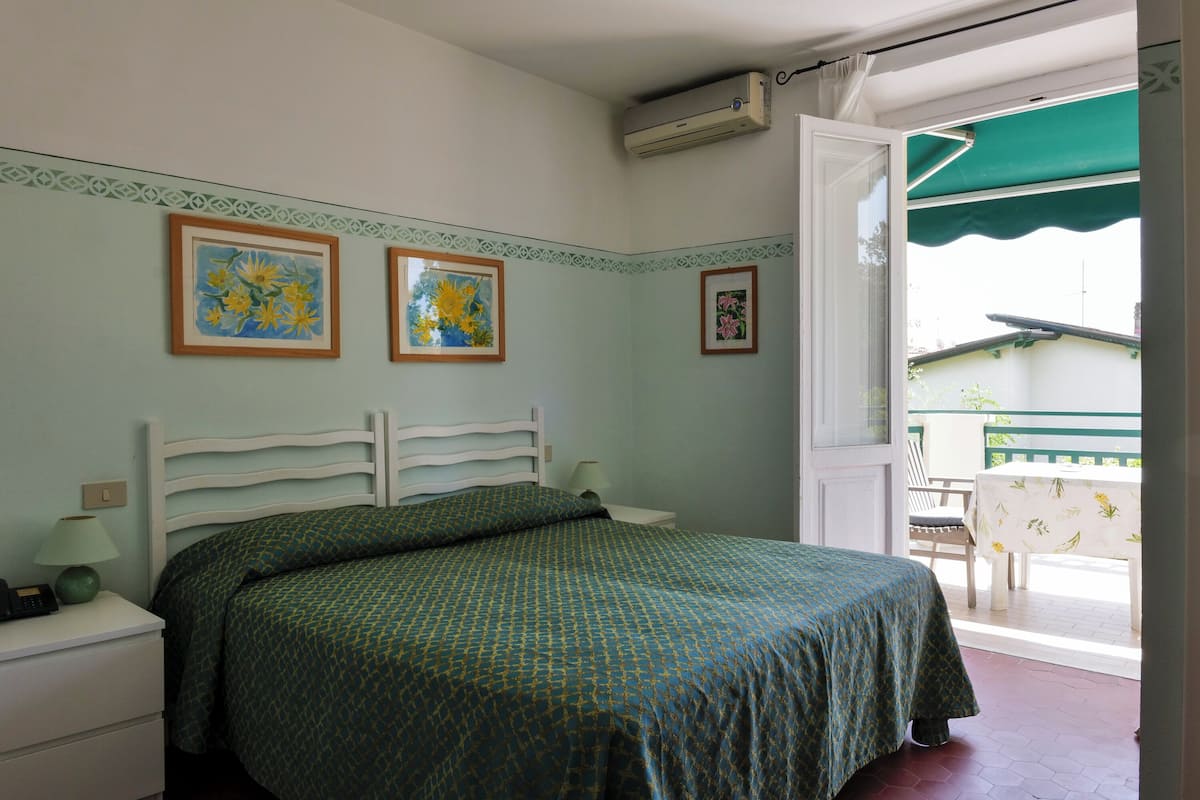 Quarto com cama, mesa de cabeceira, luminária e janela com vista para o exterior.