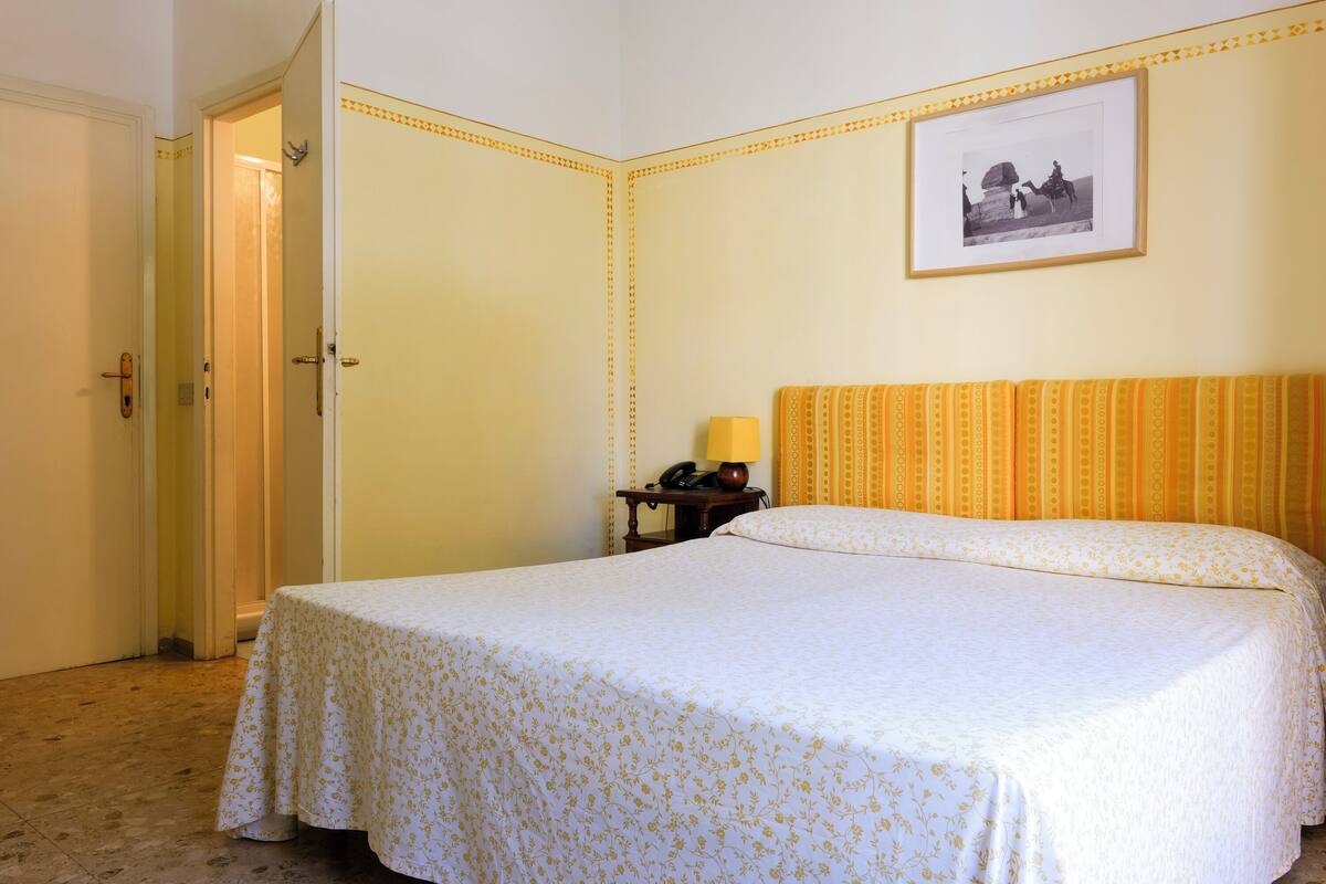 Quarto com cama, mesa de cabeceira com abajur, quadro pendurado na parede e porta que dá para outro cômodo.