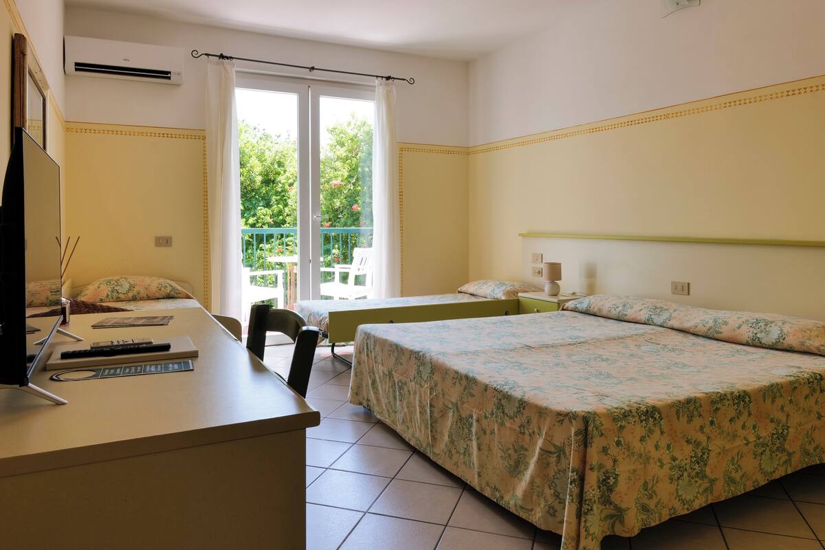 Quarto de hotel com duas camas, uma mesa com computador e varanda com vista para o verde.