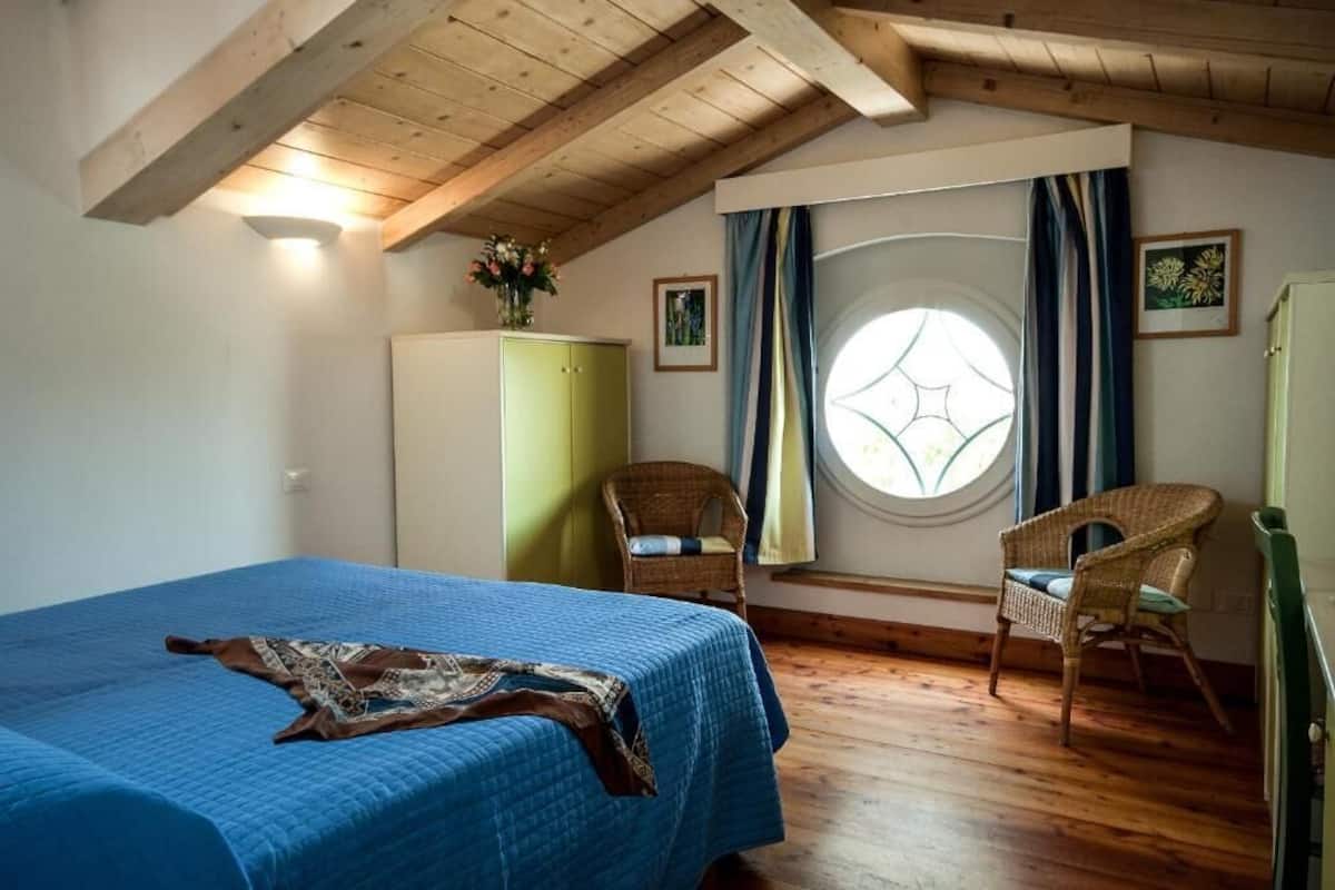 Um quarto com uma cama, duas cadeiras de vime, um guarda-roupa verde, uma janela com cortinas e um candeeiro fixado na parede.