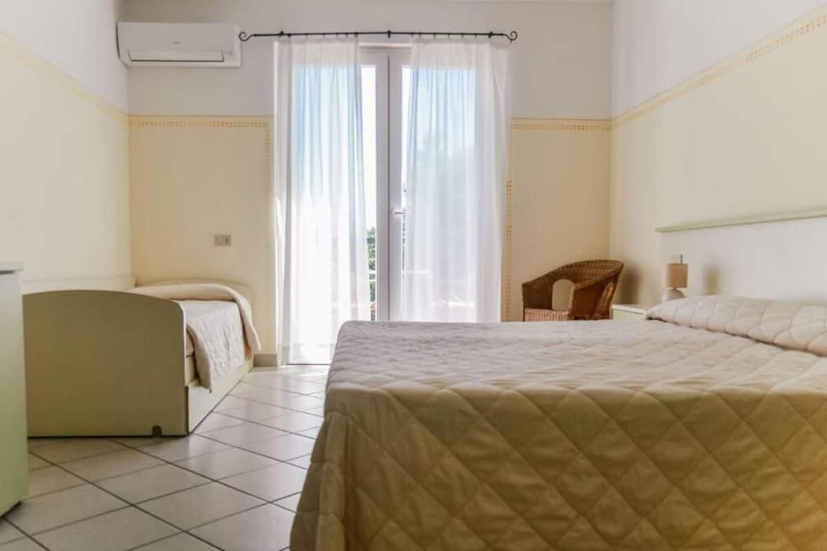 Um quarto com cama, uma cadeira, uma mesinha e uma janela com cortinas.