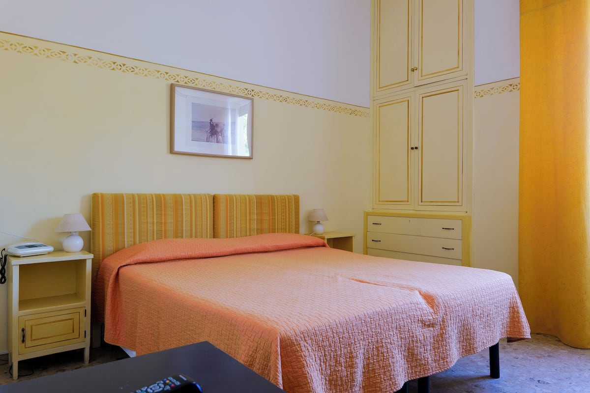 Quarto de hotel com cama, mesa de cabeceira, luminária, guarda-roupa e um quadro na parede.