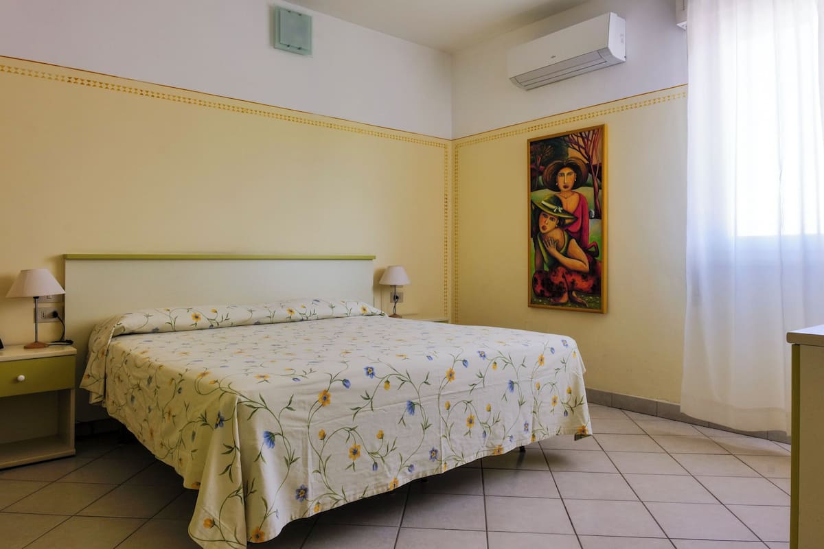 Um quarto com cama, abajures, um quadro na parede e uma janela com cortinas.