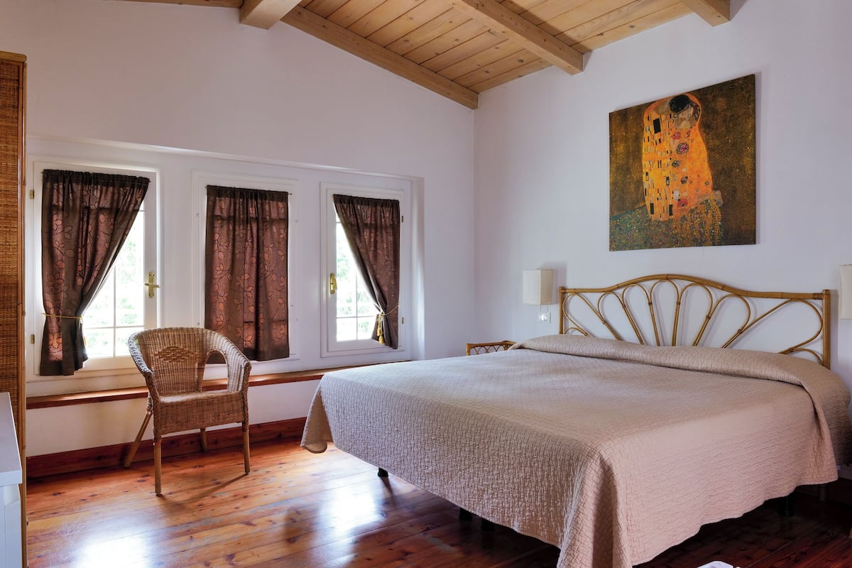 Quarto com cama, cadeira, janela com cortinas e um quadro na parede.