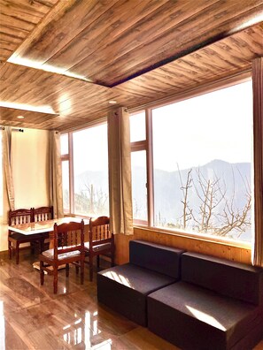 Quarto com teto de madeira, grandes janelas com vista para as montanhas, mesa de jantar de madeira com cadeiras e um sofá cinzento.
