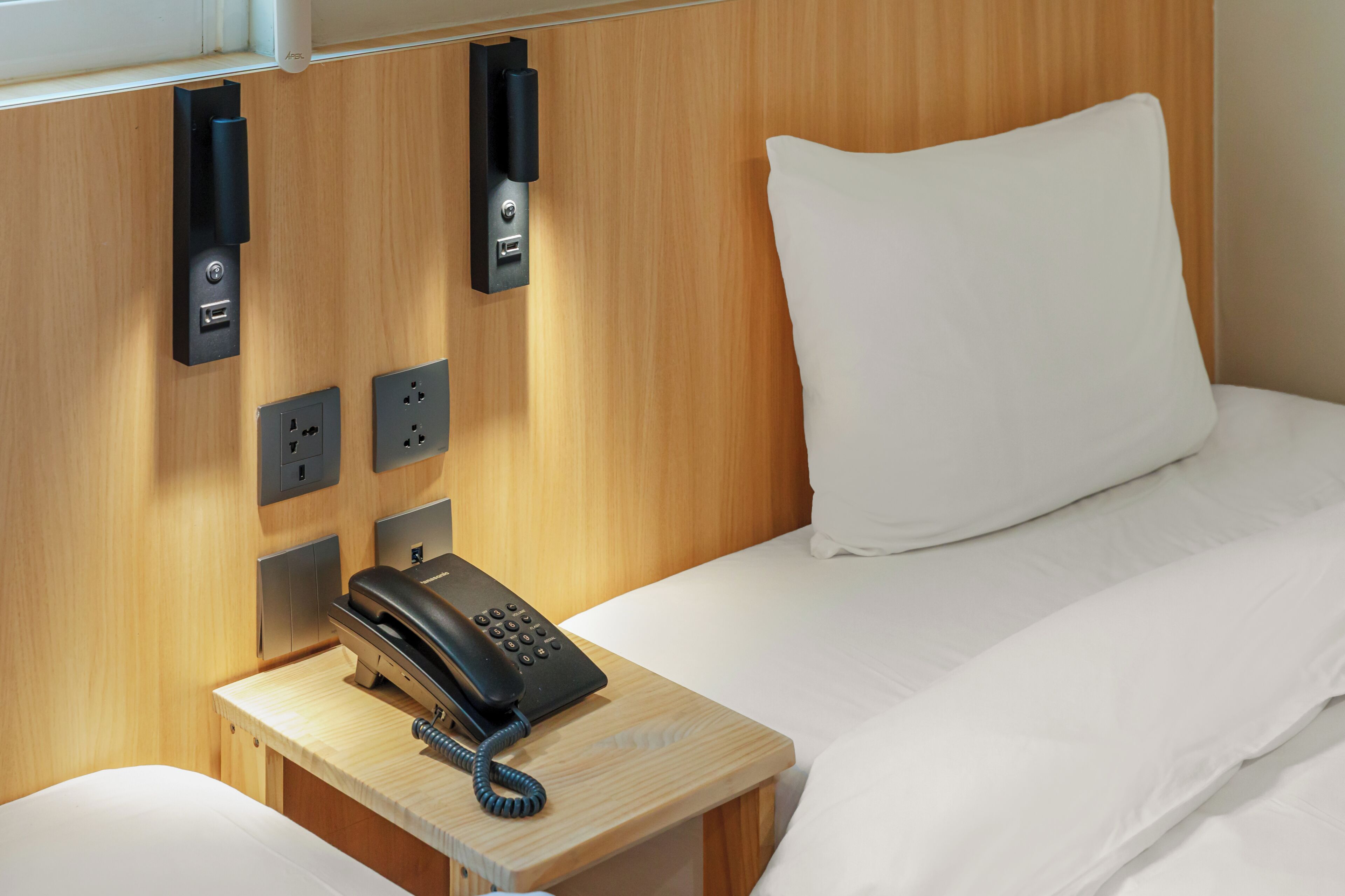 Quarto de hotel com cabeceira de madeira, telefone ao lado da cama, tomada e janela.