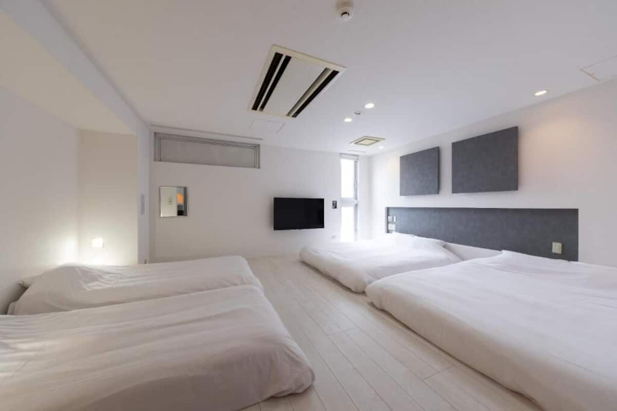Quarto de hotel moderno com três camas, televisão de tela plana na parede e uma janela grande.