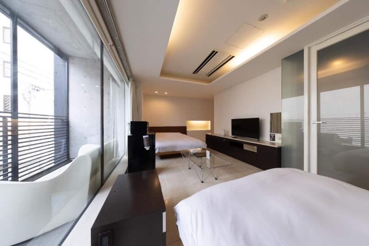 Quarto de hotel moderno com uma cama grande, uma escrivaninha, uma TV e uma varanda com uma cadeira branca.