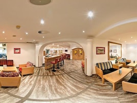 Um hall de hotel espaçoso com bar central, diversas opções de assento e vista para a área de jantar.