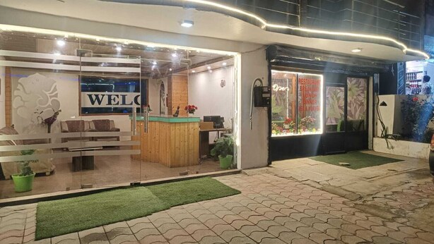 Entrada de um hotel moderno com balcão de recepção, um letreiro com a palavra 'WELCOME' e um sinal de restaurante indicando café da manhã, almoço, jantar e serviço de quarto.