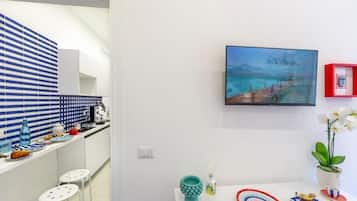 Uma cozinha moderna com painel de azulejos azul e branco, uma área de jantar com mesa posta para dois, uma TV fixada na parede e uma orquídea em vaso.