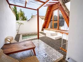 Um pequeno pátio externo com uma mesa de madeira, uma cadeira de vime, uma piscina pequena e uma estrutura de telhado de palha.