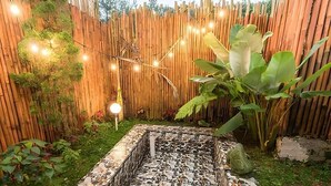 Um espelho d'água retangular com design em mosaico, cercado por uma vedação de bambu e luzes de corda.