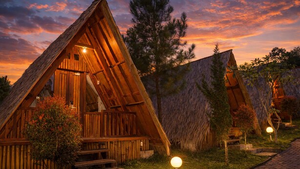 Uma cabana de madeira triangular com telhado de palha, rodeada por árvores e um caminho pavimentado.
