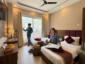 Quarto de hotel com duas camas, uma escrivaninha com abajur, uma mala e uma janela com cortinas.