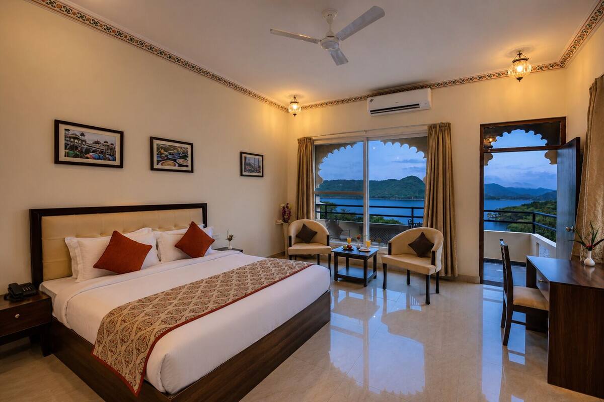 Quarto de hotel com uma cama grande, duas poltronas, uma mesa pequena e vista para um lago.