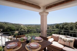 Uma varanda com uma mesa posta para refeições, um espaço de estar acolchoado e vista para uma paisagem verde.