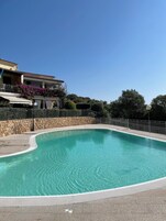 Piscina límpida com parede de pedra e grade de metal, rodeada por um edifício com varanda e flores roxas.