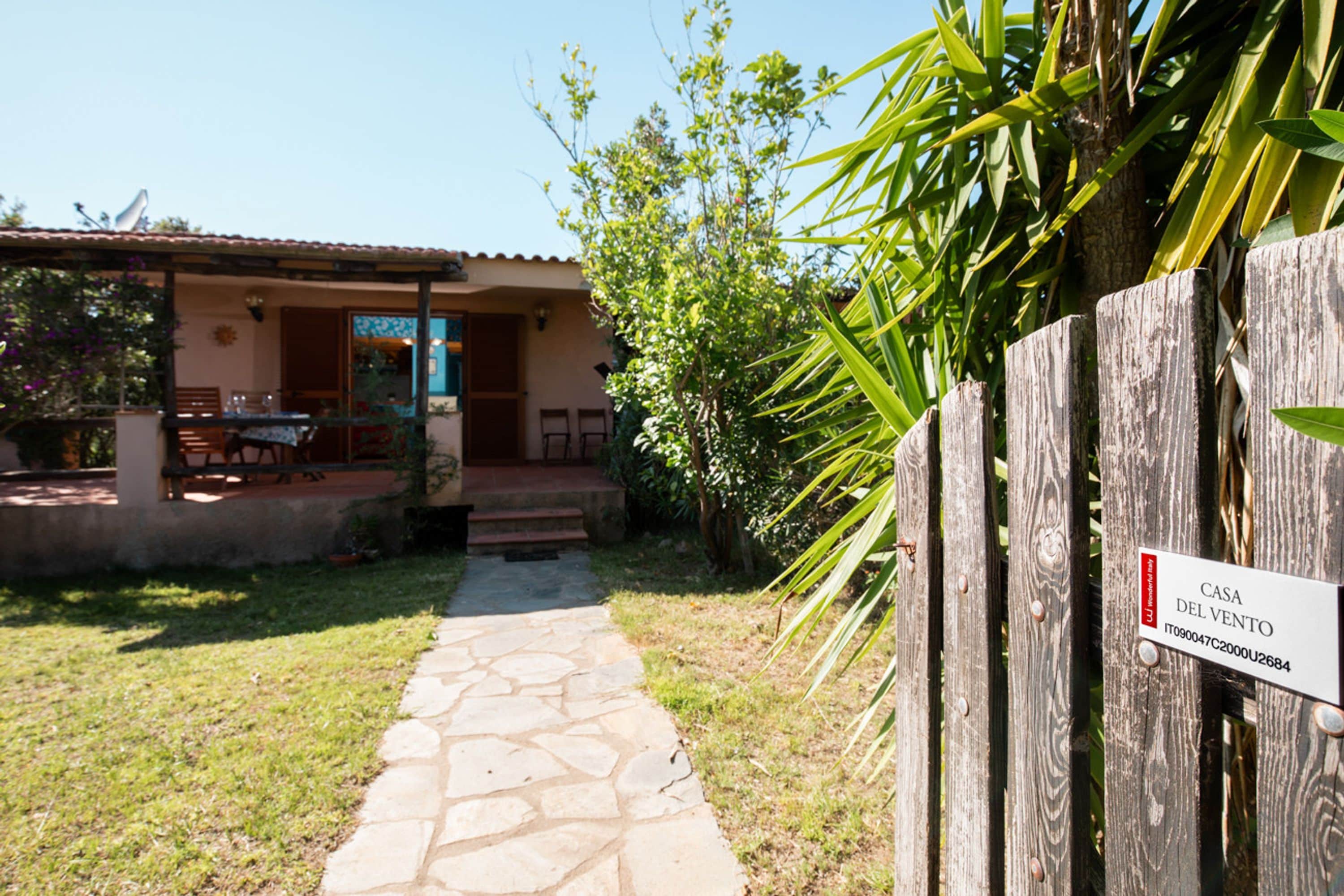 Uma trilha de pedras leva a uma casa com um pátio. Uma cerca de madeira com uma placa é visível.