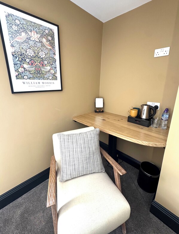 Quarto de canto com uma obra de arte emoldurada, uma mesa com chaleira, xícara e garrafa, uma cadeira e uma lixeira.