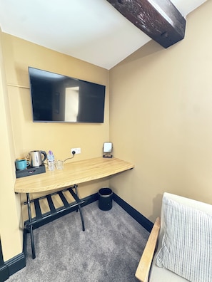 Um quarto pequeno com uma escrivaninha de madeira, uma televisão na parede, uma cafeteira, uma garrafa de água e uma lixeira.