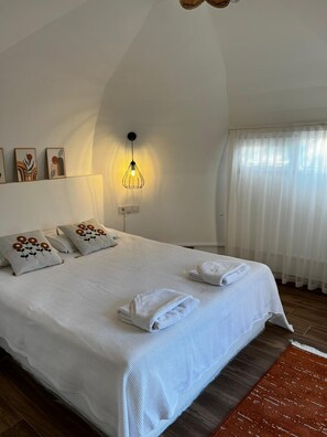 Quarto com cama grande, luminária pendente, janela com cortinas e obras de arte na parede.