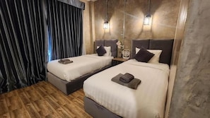 Quarto de hotel com duas camas, piso de madeira, cortina escura e luminária fixada na parede.