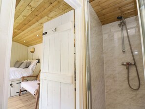 Teto de madeira, chuveiro com ducha manual, cama com travesseiros e uma mesa pequena.