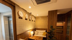 Quarto de hotel com piso de madeira, um sofá, uma mesa, uma cadeira, um vaso de plantas e um relógio de parede.