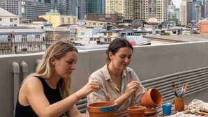 Duas pessoas sentadas à mesa, com vasos de terracota e materiais de pintura, com vista para uma paisagem urbana com edifícios altos.