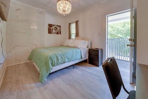 Quarto com cama, cadeira, mesa de cabeceira, candelabro, janela com persianas e porta para varanda.