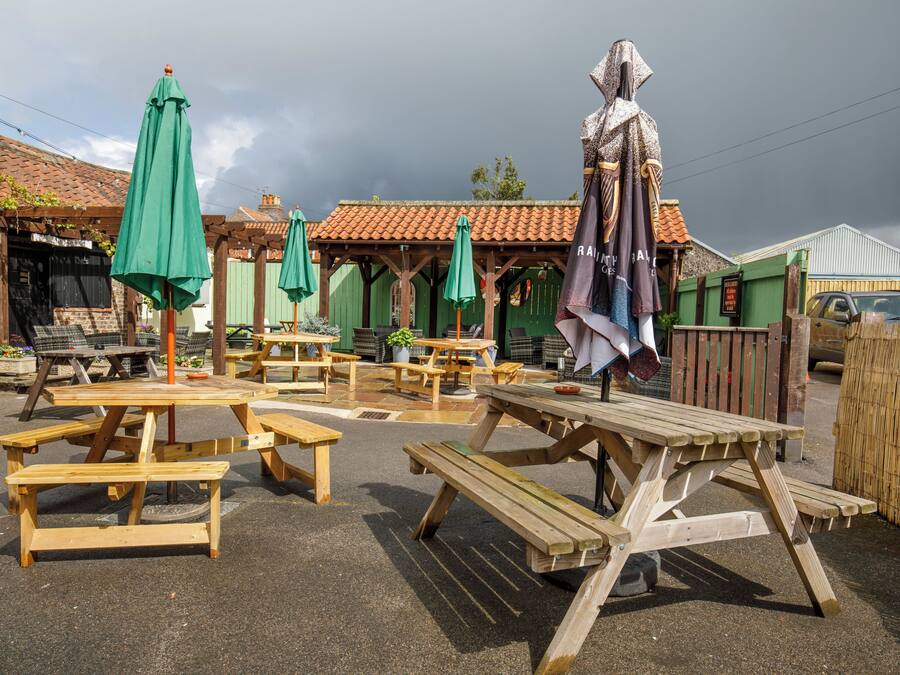 Mesas de piquenique de madeira ao ar livre, com guarda-sóis verdes, um edifício rústico com telhado de telhas e um carro estacionado nas proximidades.