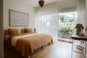 Quarto com uma cama grande, uma obra de arte na parede, uma mesa com abajur e vista para o verde lá fora.