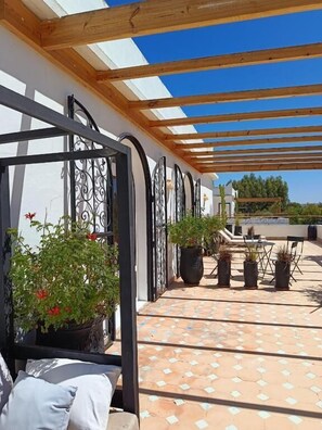 Um alpendre coberto ao ar livre com piso de azulejos, plantas em vasos e estrutura de pérgola de madeira.