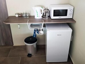 Uma cozinha compacta equipada com micro-ondas, um pequeno frigorífico, um cesto de lixo e uma prateleira com utensílios de cozinha.