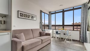 Uma sala de estar moderna com sofá, mesa de jantar e cadeiras, área de cozinha e janelas amplas com cortinas.