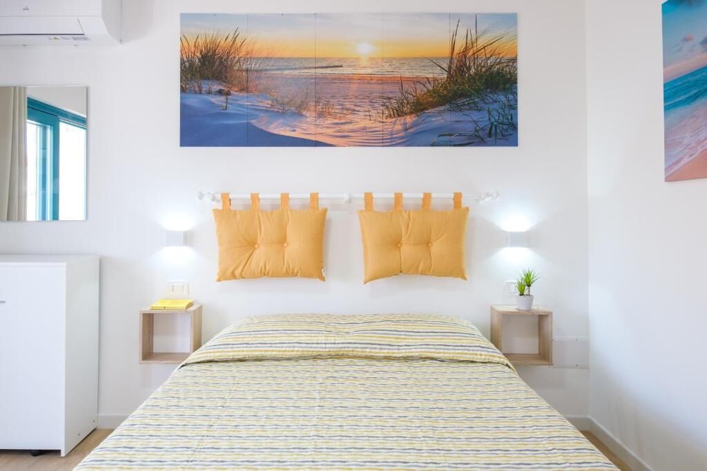 Um quarto com uma cama, duas mesas de cabeceira, um criado-mudo, uma luminária na parede e um quadro sobre a cama.