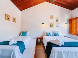 Quarto com duas camas, cada uma com roupa de cama branca e detalhes em azul-turquesa, uma pequena mesa de cabeceira de madeira e arte nas paredes.