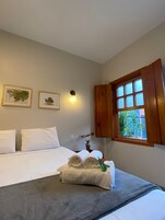 Cama bem arrumada com lençóis brancos, um cobertor cinza e um vaso com flores sobre a cama. Há duas molduras com quadros na parede, uma luminária fixada na parede e uma janela com persianas de madeira.